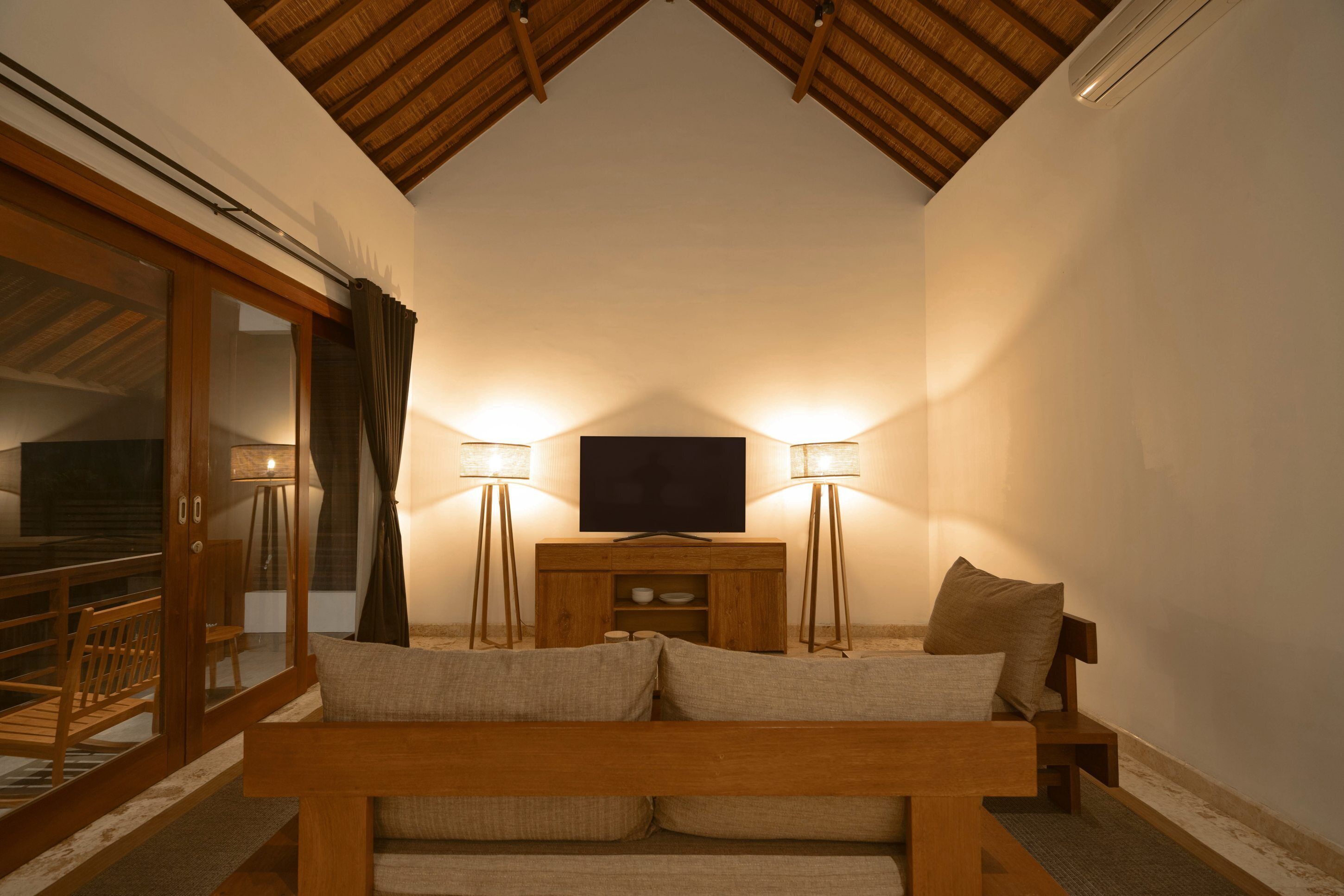 Um quarto aconchegante com teto de madeira, televisão, sofá e duas luminárias de chão.