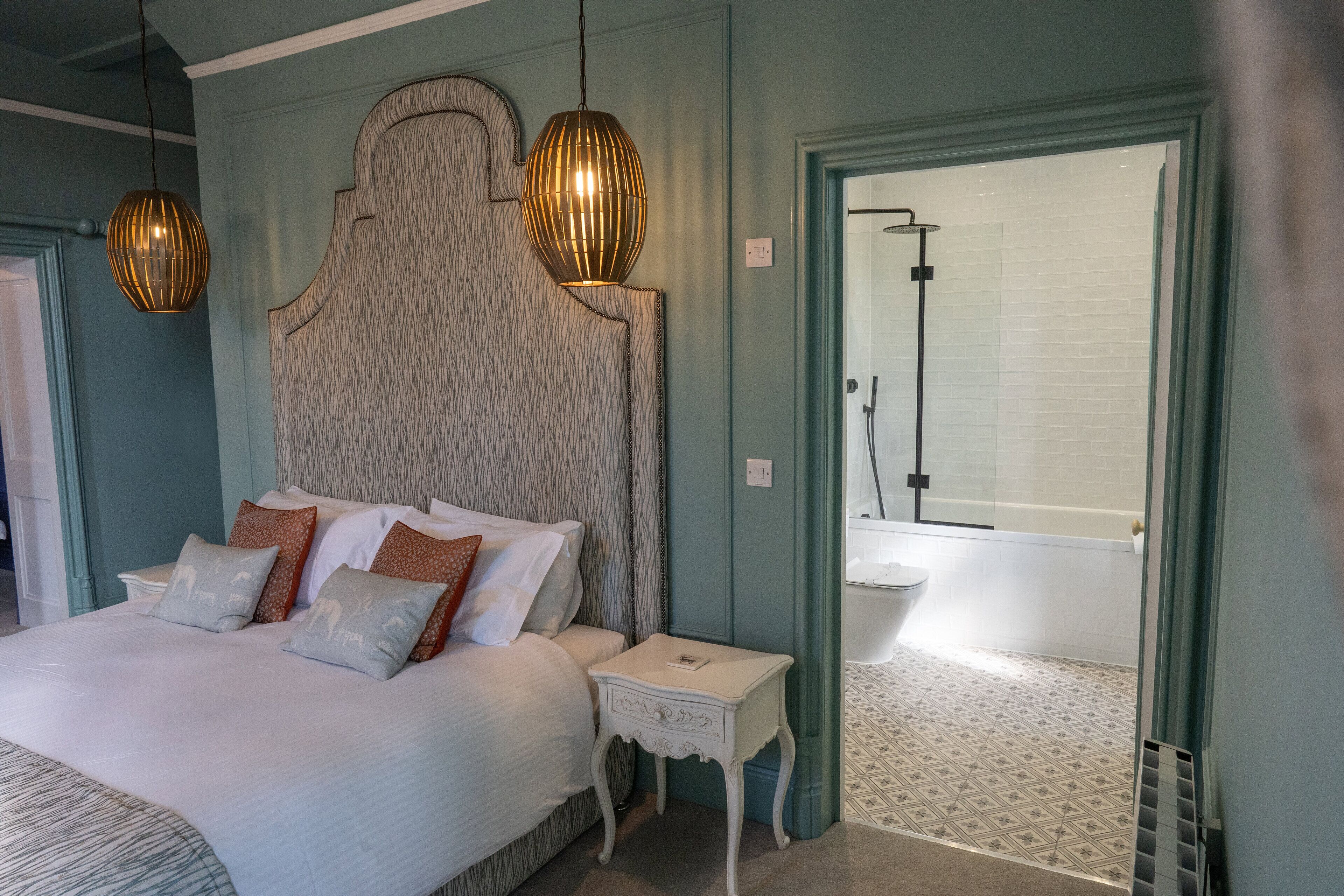 Quarto com cama grande, mesas de cabeceira, luminária pendente e vista para um banheiro com banheira e chuveiro.