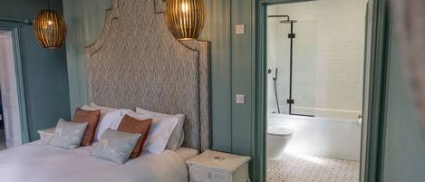 Quarto com cama grande, mesas de cabeceira, luminária pendente e vista para um banheiro com banheira e chuveiro.
