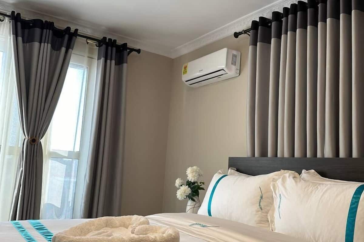 Quarto de hotel com cama, janela com cortinas, ar condicionado e mesa de cabeceira com um vaso de flores.