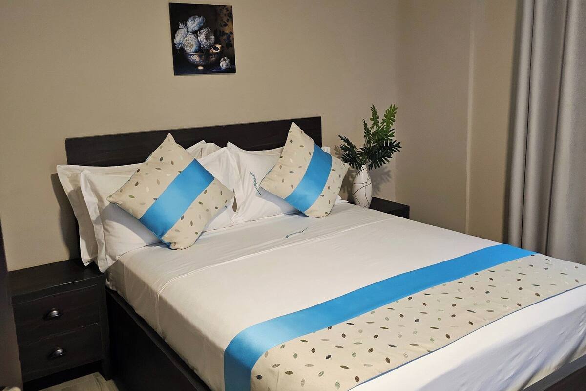 Cama bem arrumada com detalhe azul, cabeceira de madeira escura, mesa de cabeceira pequena e um quadro fixado na parede.