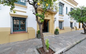 Um edifício com entrada de pedra, uma árvore com frutos amarelos e um pequeno vaso com arbustos verdes.