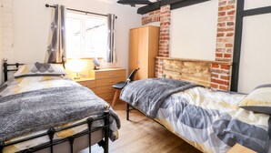 Um quarto com duas camas de solteiro, um guarda-roupa de madeira, uma mesa de trabalho com uma luminária e uma janela com cortinas.