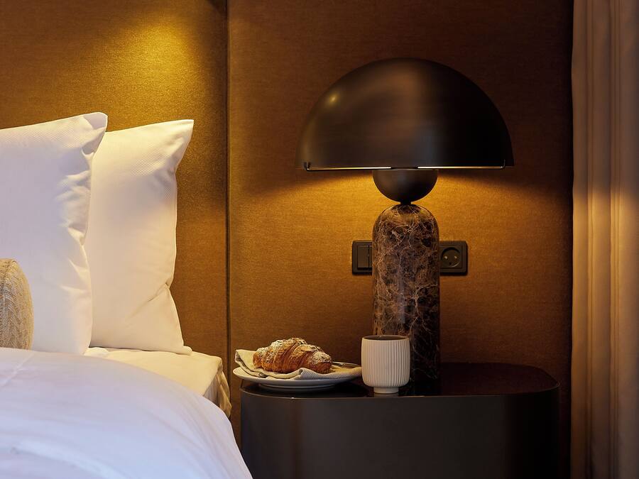 Cama bem arrumada com lençóis e almofadas brancas, abajur com base de mármore e um prato com um croissant em uma mesinha ao lado da cama.