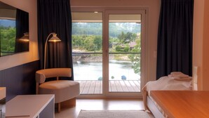 Quarto de hotel com cama, poltrona, escrivaninha, televisão e vista para um rio e vegetação.