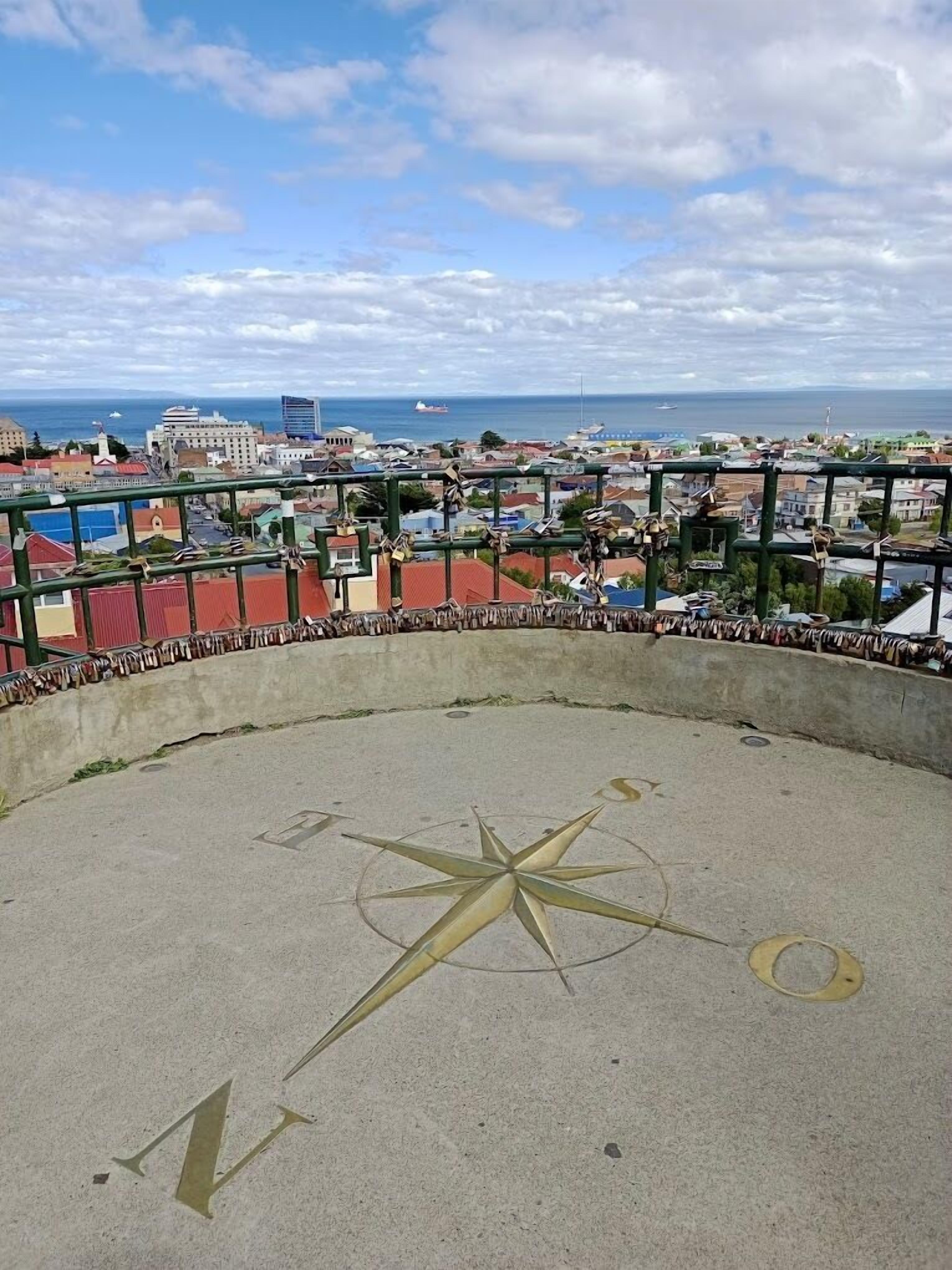 Paisagem costeira urbana com uma rosa dos ventos em destaque no chão de concreto, com vista para um porto com navios e edifícios.