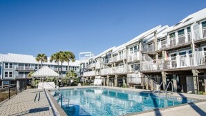 Área de piscina com piscina azul translúcida, espaço coberto para sentar e um edifício com varandas.