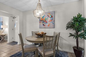 Uma sala de jantar com uma mesa redonda, cadeiras de madeira, um candelabro, um quadro abstrato e colorido e uma planta em vaso.