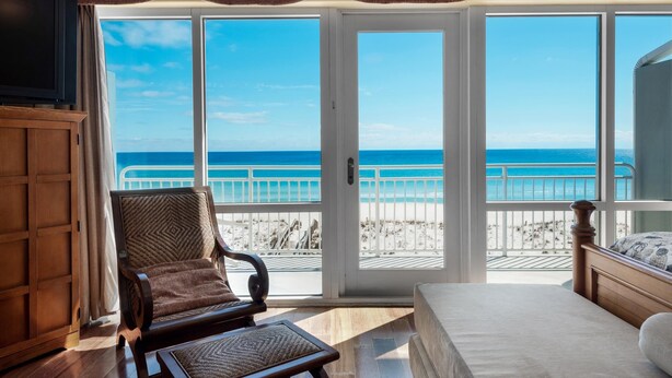 Quarto com cama, cadeira e televisão. Possui janelas amplas com vista para a praia e oceano.