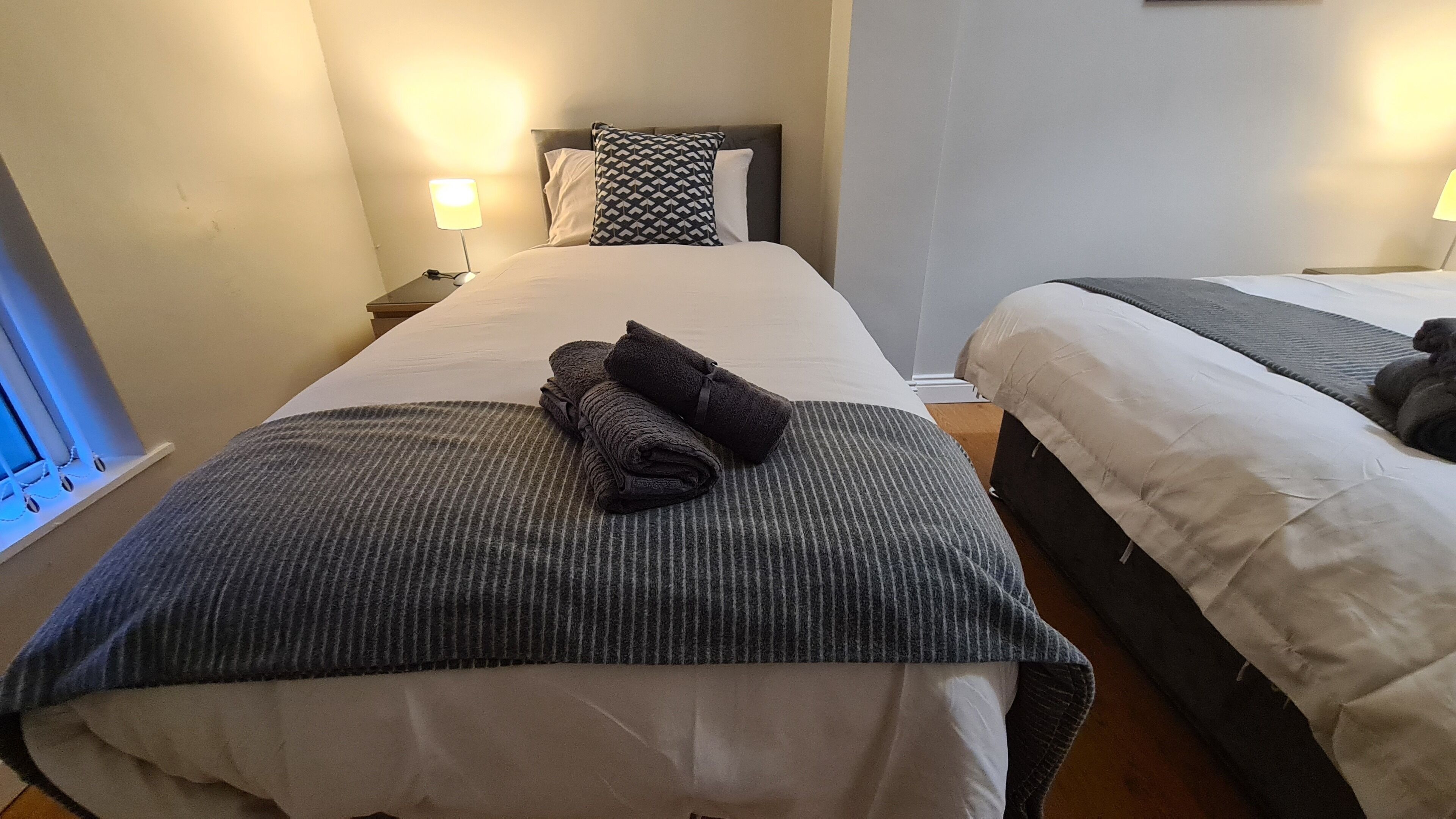 Quarto de hotel com uma cama grande, uma cama menor e uma mesa de cabeceira com abajur.