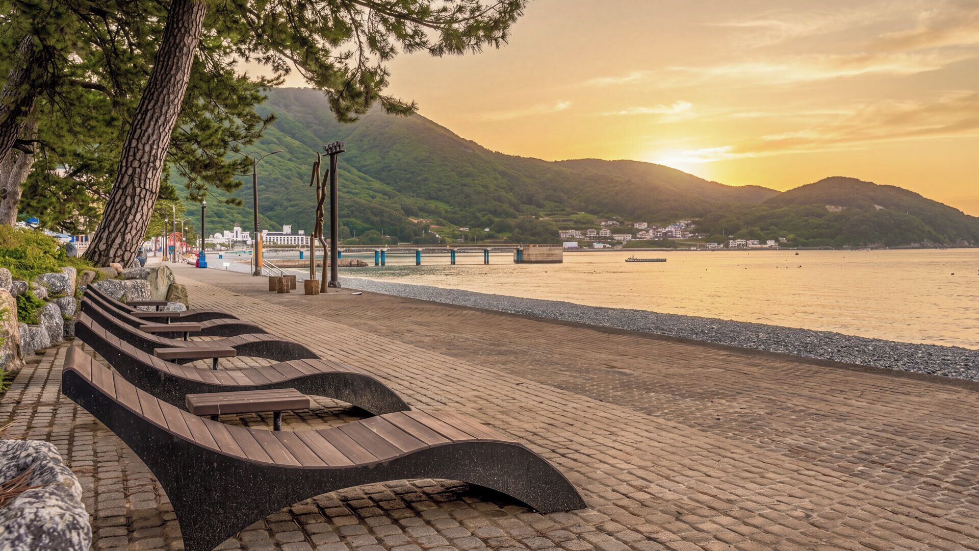 Um calçadão à beira-mar com bancos, uma ponte e colinas ao fundo.