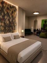 Quarto de hotel moderno com uma cama grande, parede de detalhes em pedra, sofá verde, escrivaninha com luminária e um quadro.