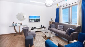 Uma sala de estar moderna com um sofá cinzento, poltronas azuis, uma mesa de centro de vidro, uma pequena mesa lateral, uma TV fixada na parede e uma grande janela com cortinas.