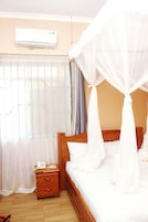 Quarto com cama de madeira, roupa de cama branca, mesa de cabeceira com telefone e janela com cortinas leves.