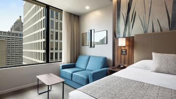 Quarto de hotel com uma cama, um sofá, uma mesinha e uma janela ampla com vista para a cidade.