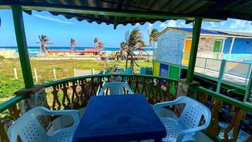 Uma área de estar coberta ao ar livre, com mesa e cadeiras, com vista para uma praia com palmeiras e edifícios ao fundo.