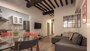 Uma sala de estar moderna com uma mesa de jantar de vidro, cadeiras vermelhas, um sofá cinzento e uma televisão fixada na parede.
