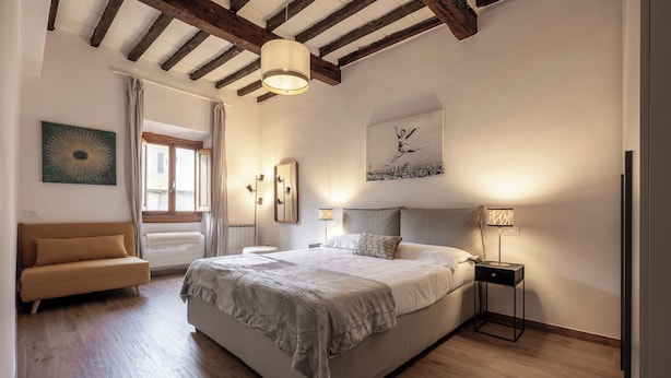Um quarto com uma cama grande, uma mesinha de cabeceira, um abajur, uma janela com cortinas, um sofá e um quadro na parede.