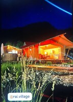 Uma área de refeições ao ar livre bem iluminada, com um toldo vermelho, cercada por uma cerca de madeira rústica e um caminho de pedra.