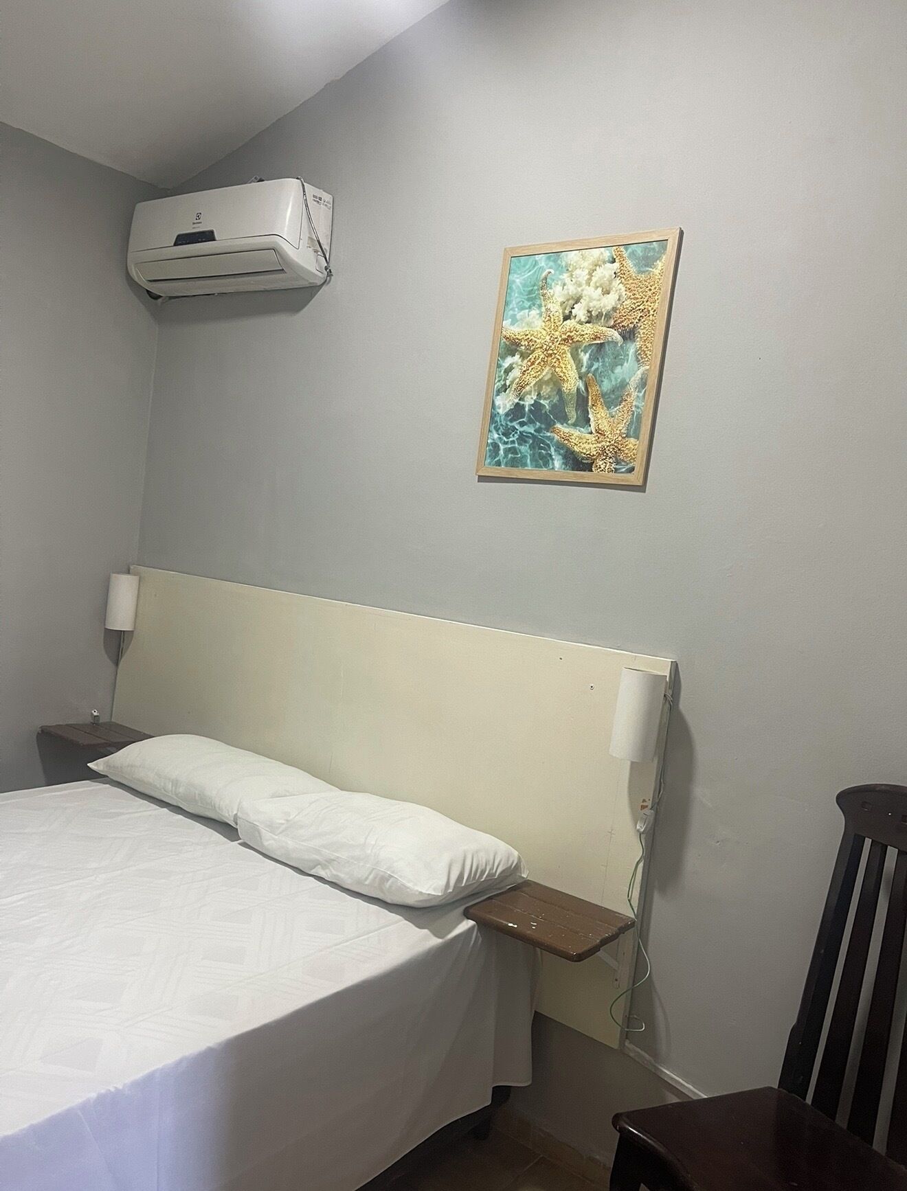 Quarto de hotel com uma cama, mesa de cabeceira, ar condicionado fixado na parede e uma fotografia emoldurada de uma estrela-do-mar.