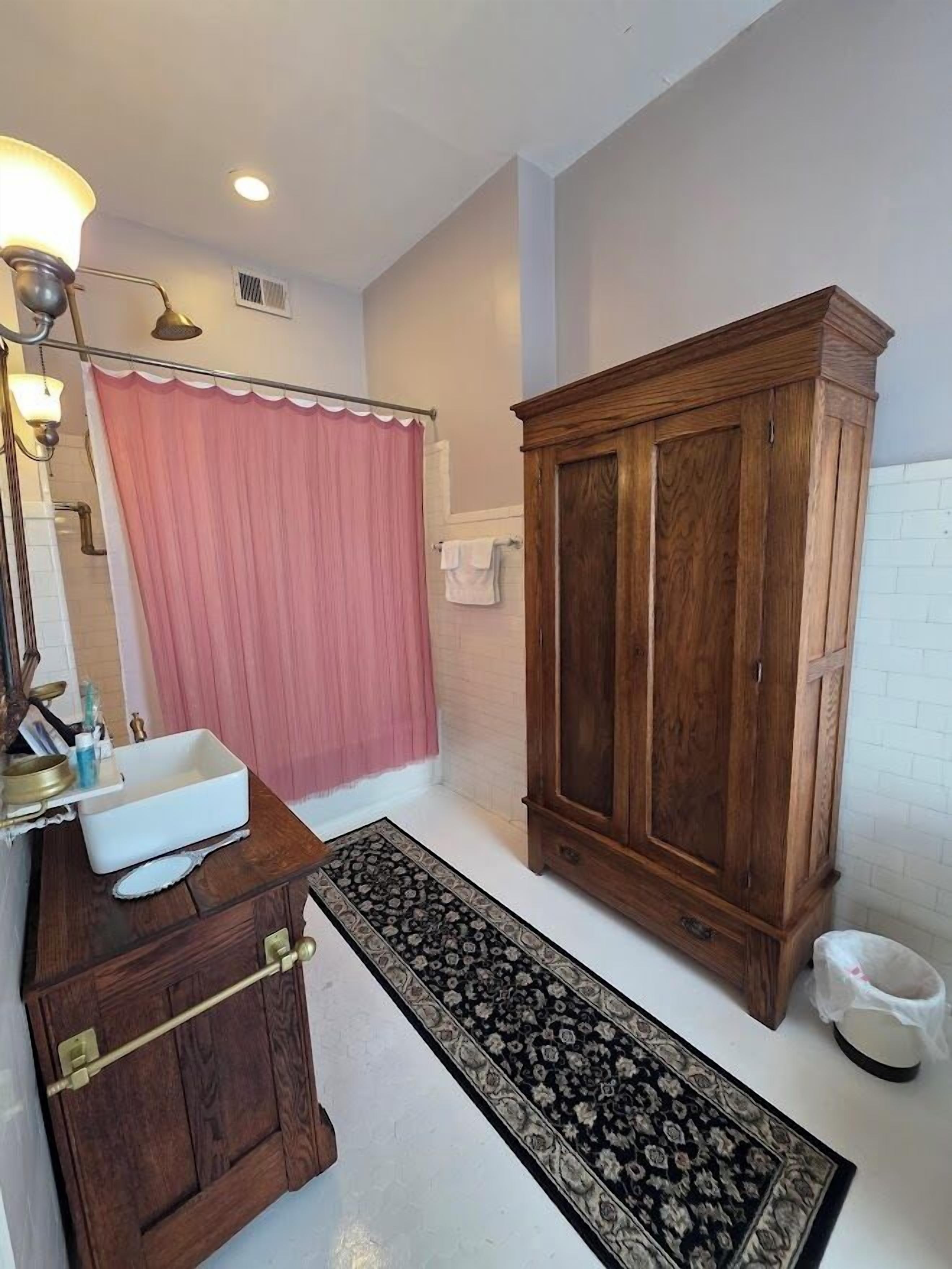 Casa de banho com armário de madeira, pia branca, cortina de chuveiro rosa e tapete estampado.