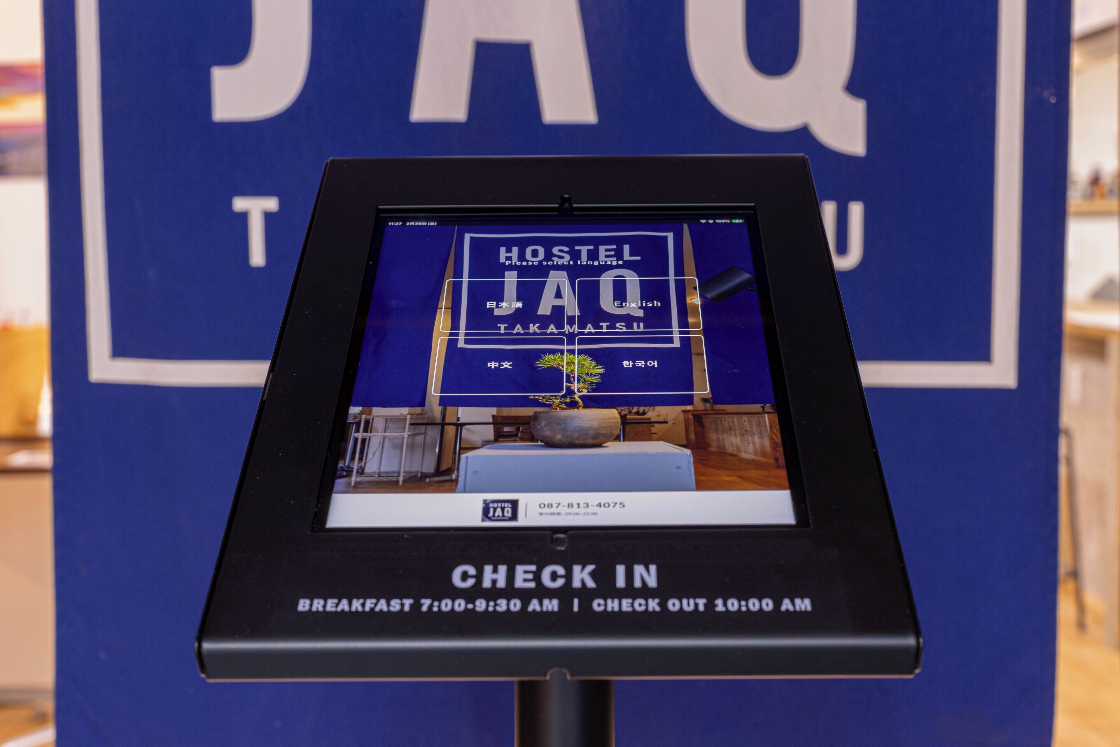 Quiosque de check-in digital com tela exibindo um anúncio de hostel, incluindo número de contato e horário do café da manhã.