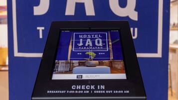 Quiosque de check-in digital com tela exibindo um anúncio de hostel, incluindo número de contato e horário do café da manhã.
