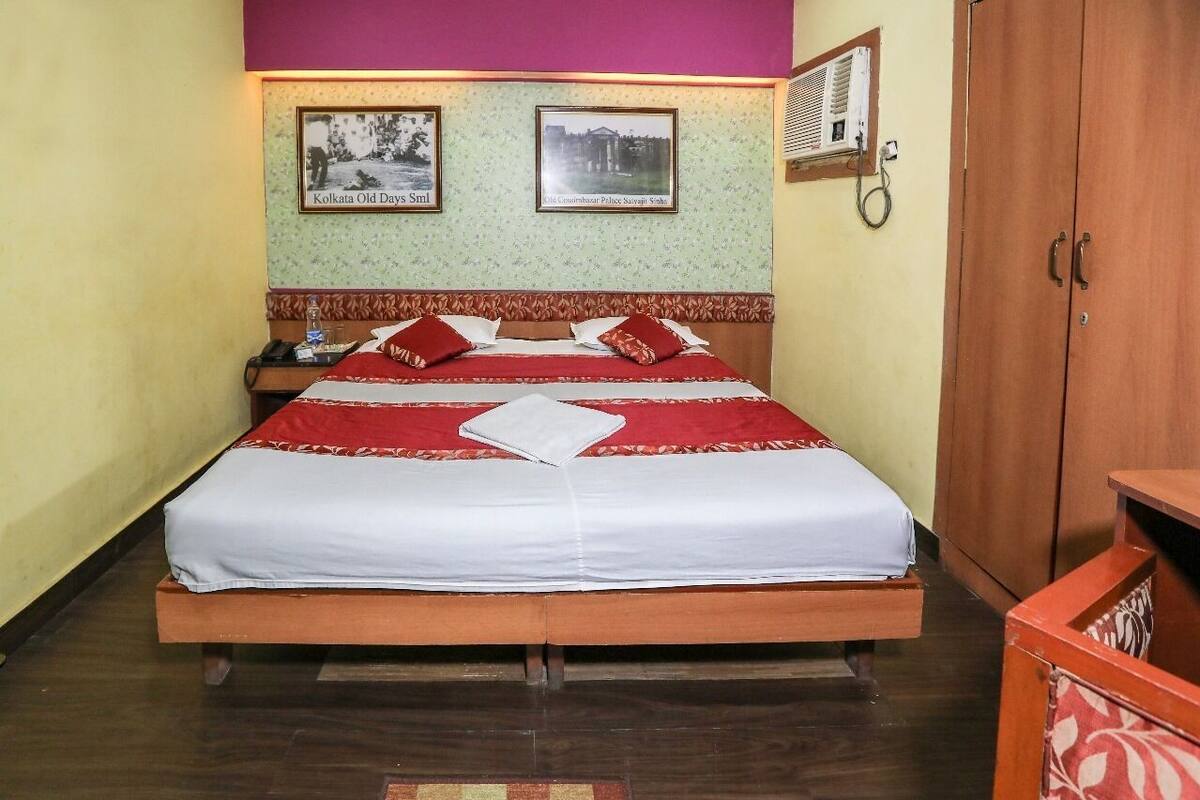 Uma cama de solteiro com estrutura de madeira, edredom branco e vermelho, e duas almofadas. Há uma mesa de cabeceira com um abajur e uma garrafa de água. O quarto possui um guarda-roupa de madeira e um ar-condicionado fixado na parede.