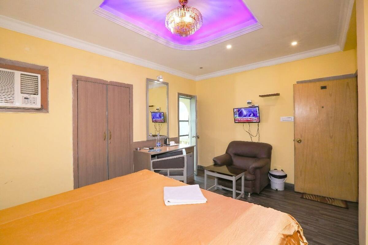 Quarto de hotel com cama, escrivaninha, cadeira, televisão, guarda-roupa e luz no teto.