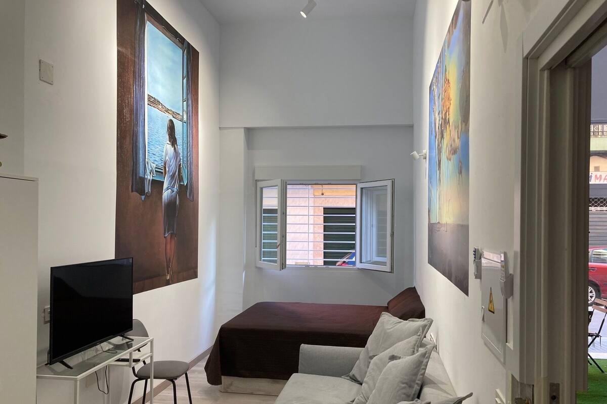 Quarto de hotel pequeno, com uma cama, televisão, escrivaninha e janela com persianas.
