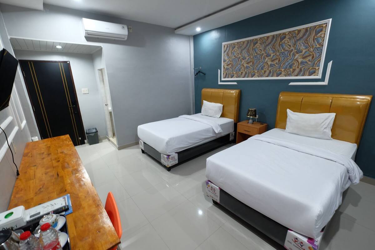 Quarto de hotel com duas camas, uma escrivaninha de madeira, uma televisão e ar condicionado fixado na parede.