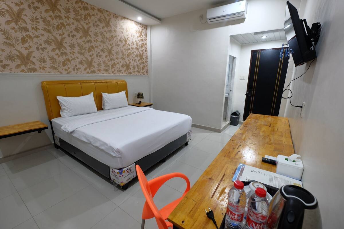 Quarto de hotel com cama, uma mesa de madeira com cadeira, TV fixada na parede e aparelho de ar condicionado.
