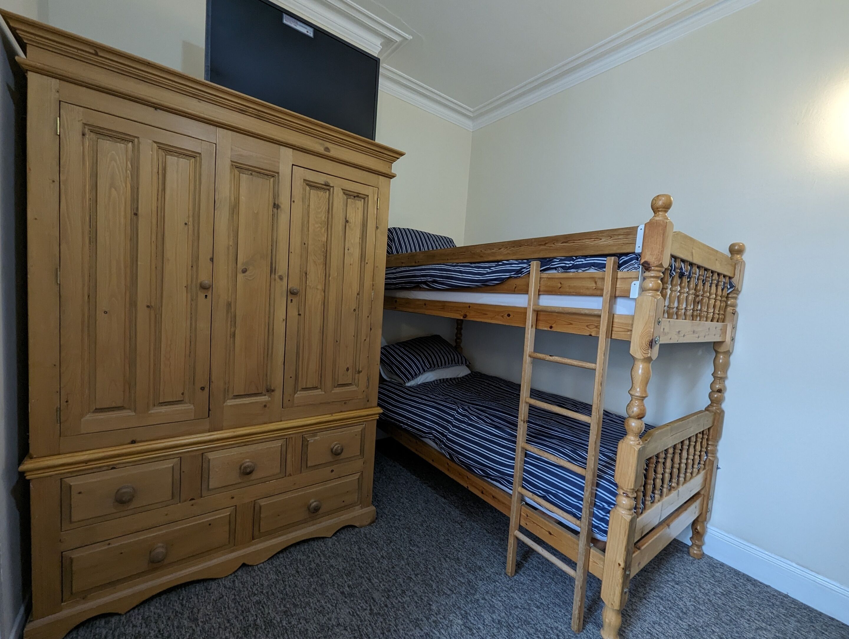 Uma cama beliche com escada de madeira, um guarda-roupa de madeira com várias gavetas e um painel preto acima do guarda-roupa.