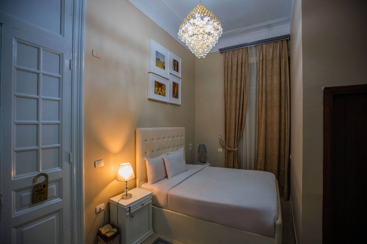 Quarto de hotel com cama, mesa de cabeceira, abajur, candelabro e uma porta.
