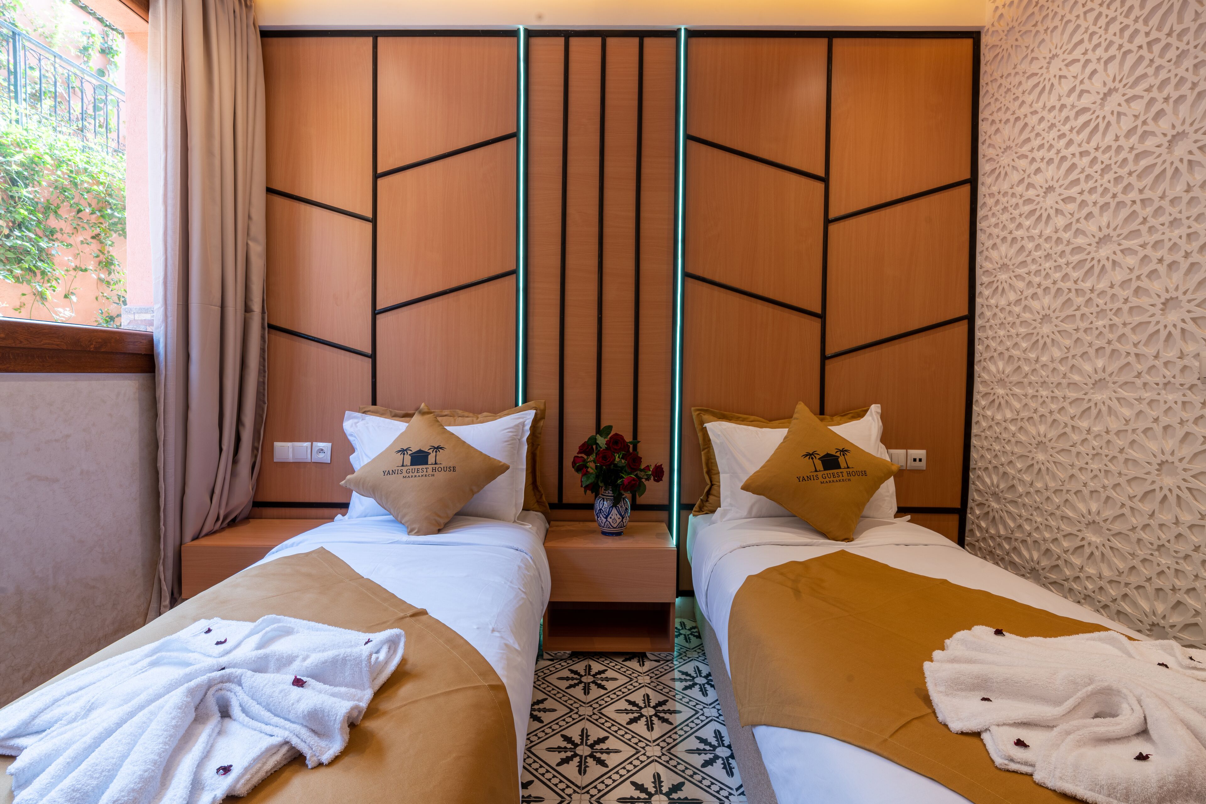 Quarto de hotel com duas camas, cada uma com cabeceira marrom, roupa de cama branca e uma toalha na cama. Há uma mesinha entre as camas com um vaso de flores.