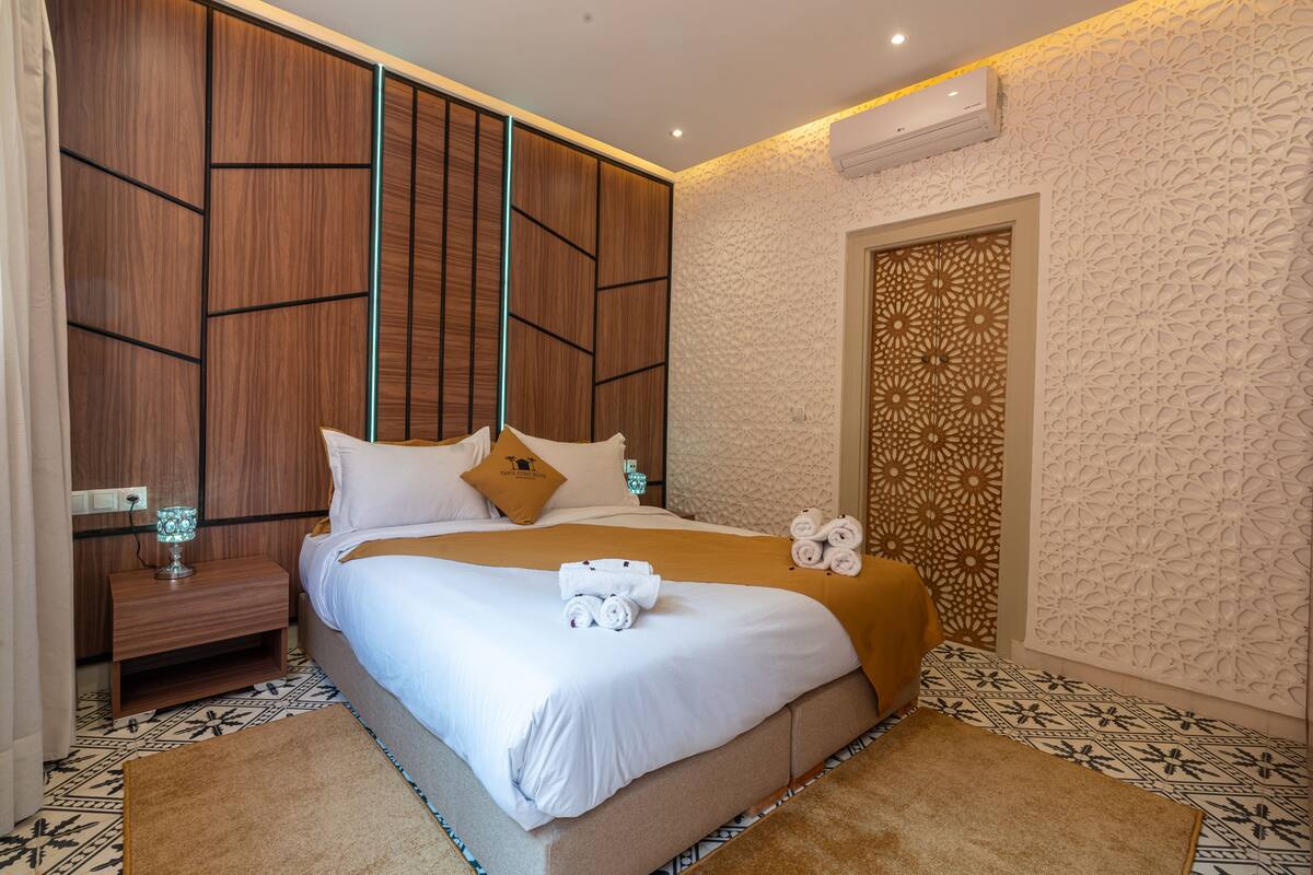 Quarto bem arrumado com uma cama grande, uma mesinha de cabeceira, ar condicionado fixado na parede e um painel decorativo.