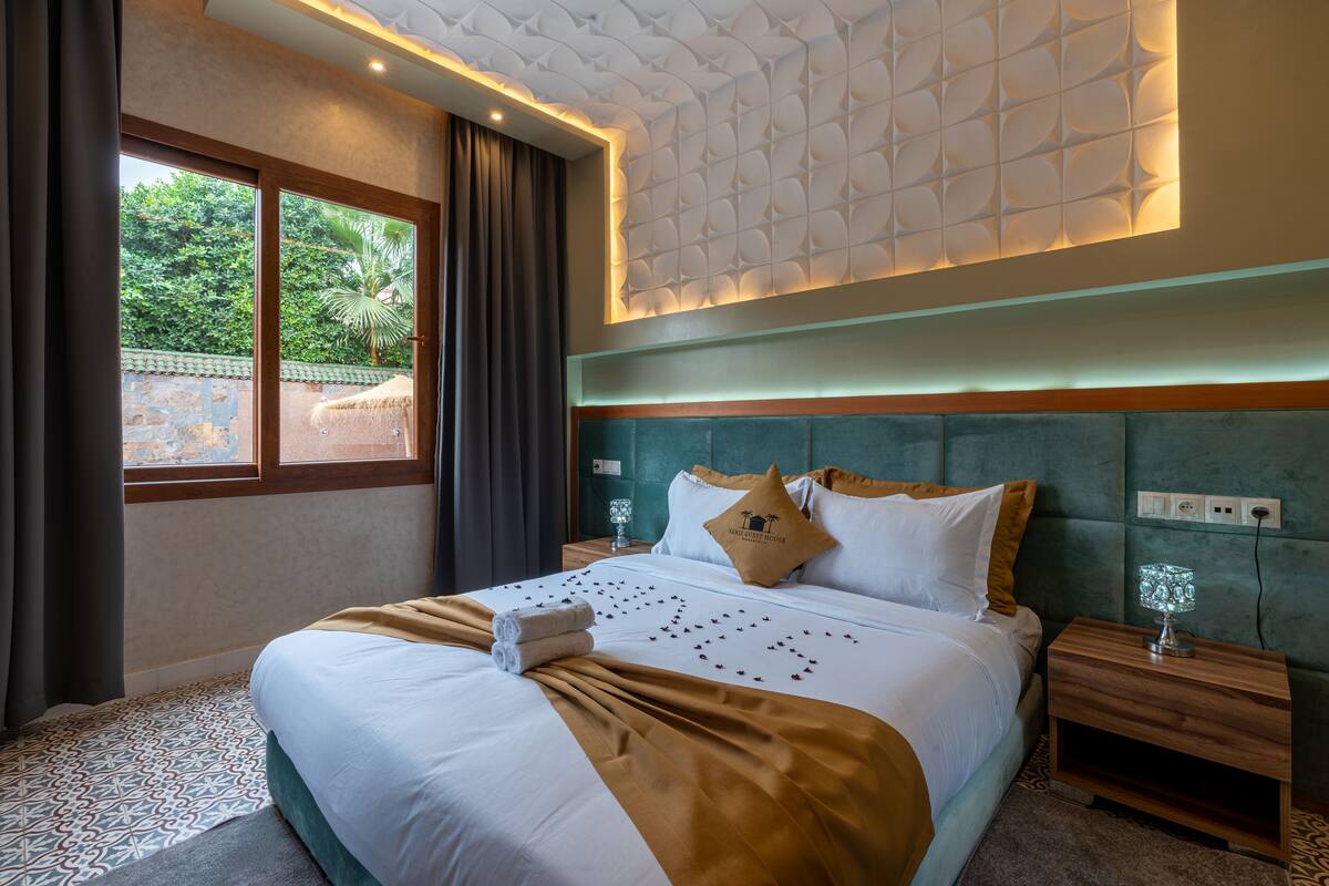 Quarto de hotel com cama grande, mesa de cabeceira, luminária na parede e janela com cortinas.