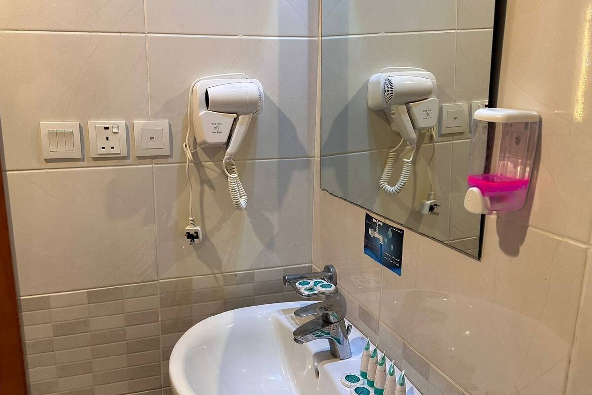 Casa de banho com lavatório, dois secadores de cabelo fixados na parede, espelho e dispensador de sabonete.