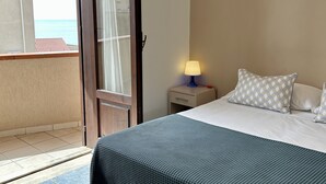 Quarto de hotel com cama, mesa de cabeceira com luminária, janela com cortinas e vista para o mar.