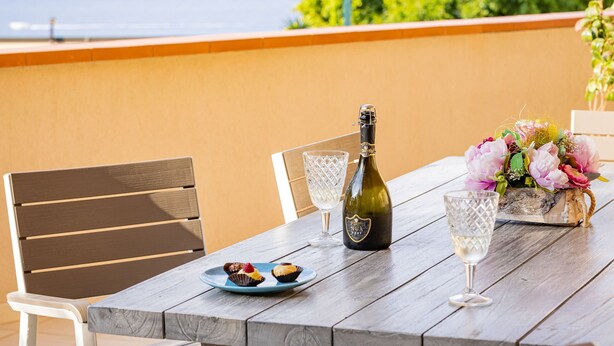 Uma mesa de madeira com uma garrafa, copos e um prato de comida num pátio exterior com vista para o mar.