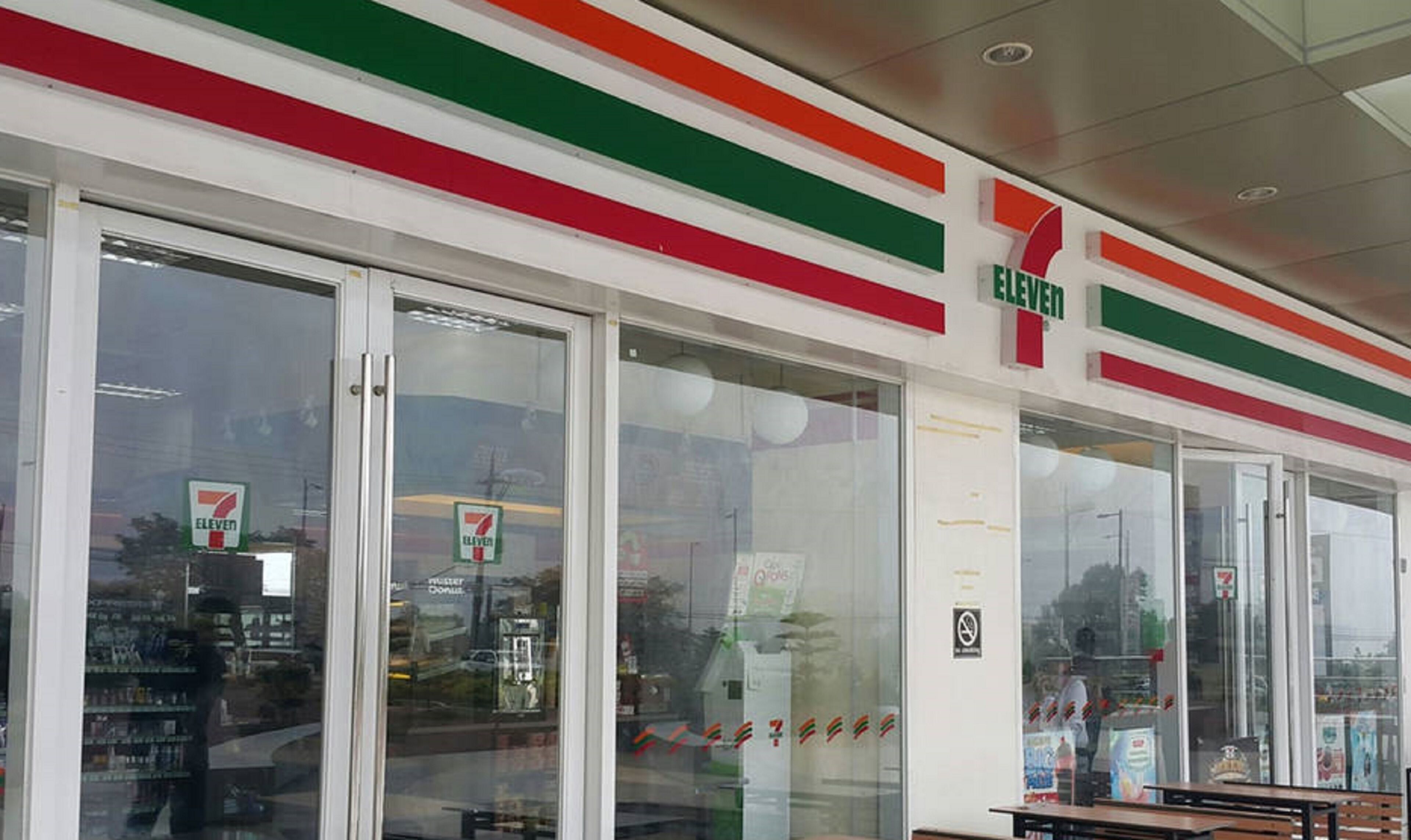 Uma loja 7-Eleven com grandes portas de vidro, toldo listrado em vermelho e verde, e placa de proibido fumar na parede.
