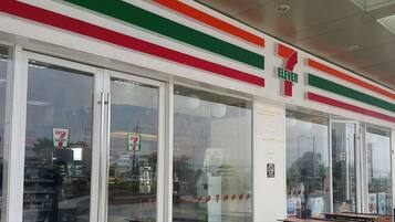 Uma loja 7-Eleven com grandes portas de vidro, toldo listrado em vermelho e verde, e placa de proibido fumar na parede.