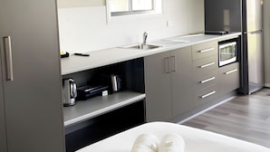 Cozinha moderna com armários cinza, uma pia de aço inoxidável e um forno embutido. Uma cama com roupa de cama branca em primeiro plano.