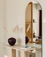 Um espelho reflete um quarto com uma mesa de madeira, um vaso preto com plumas de capim-do-campo e um livro.
