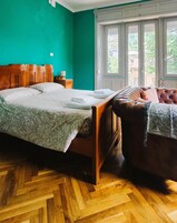 Quarto com cama e mesa de cabeceira de madeira, janela com vista para áreas verdes e uma poltrona de couro marrom.