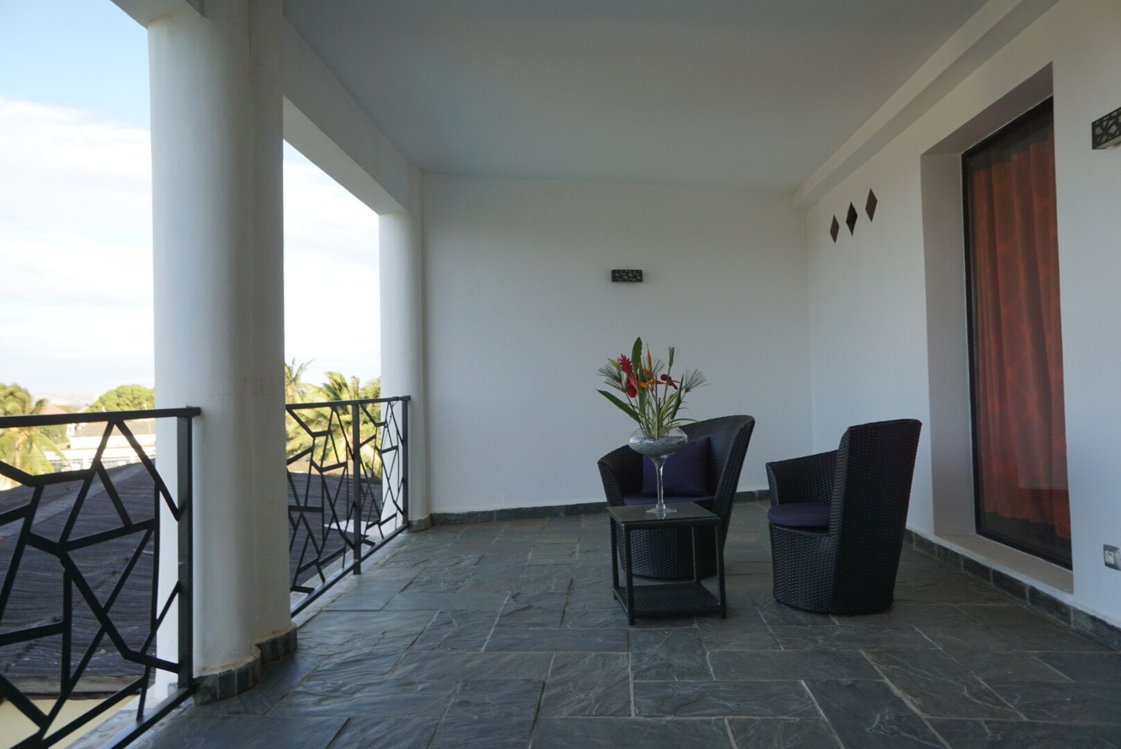 Uma varanda com duas cadeiras de vime, uma mesa pequena com um vaso de flores e vista para o mar.