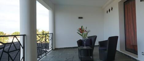 Uma varanda com duas cadeiras de vime, uma mesa pequena com um vaso de flores e vista para o mar.