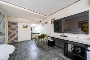 Quarto de hotel compacto com uma cama, uma escrivaninha com cadeira, televisão e área de cozinha compacta.
