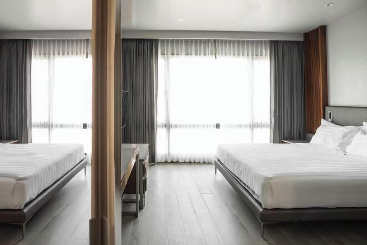 Quarto de hotel com duas camas, uma escrivaninha e amplas janelas com cortinas.