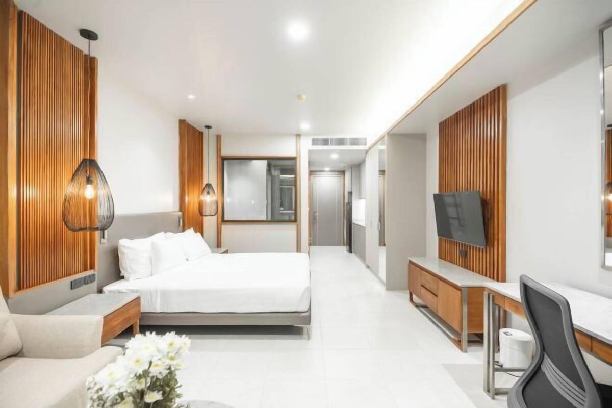 Quarto de hotel moderno com uma cama, escrivaninha, cadeira, televisão e banheiro ao fundo.